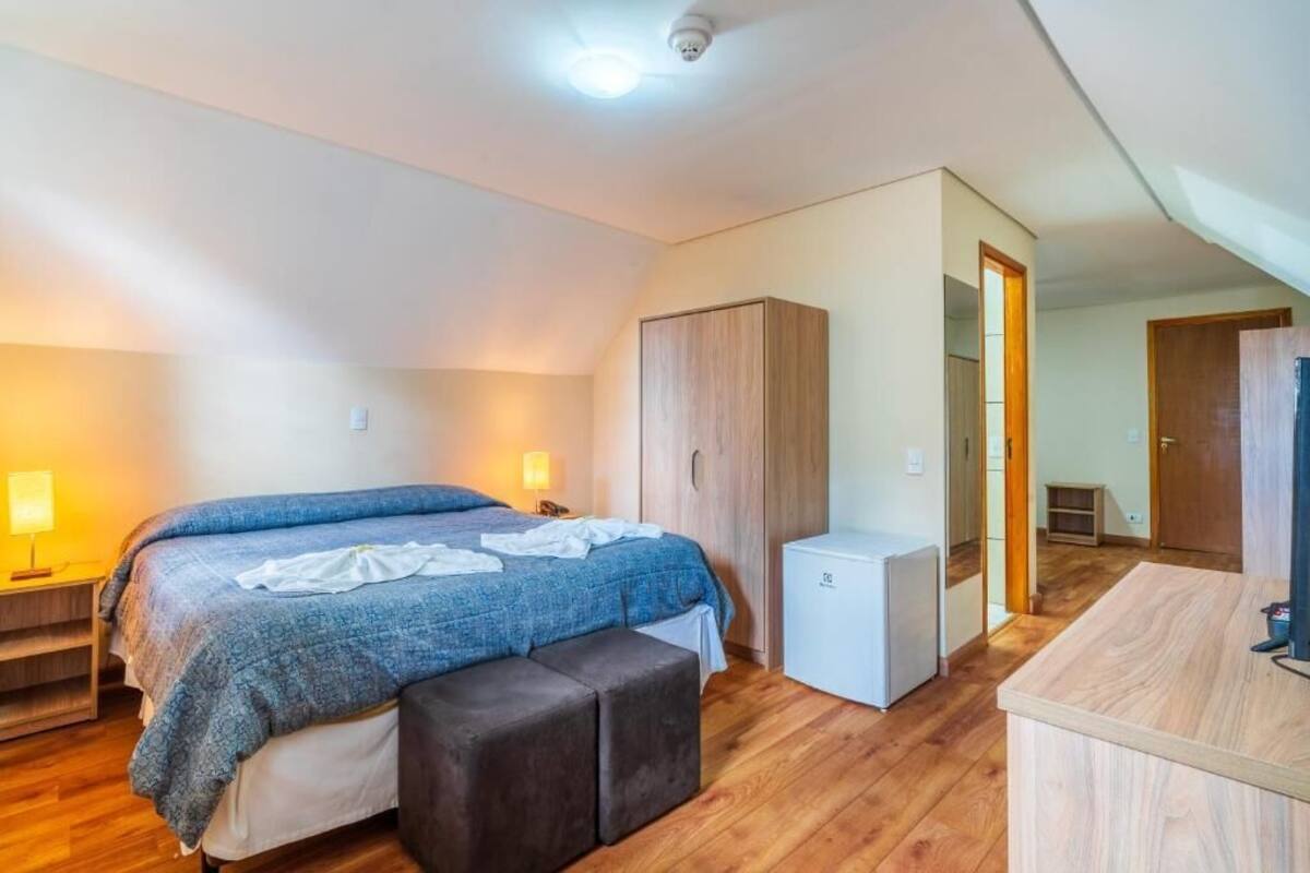 Quarto com cama, luminárias de cabeceira, guarda-roupa, mini-frigorífico e escrivaninha.