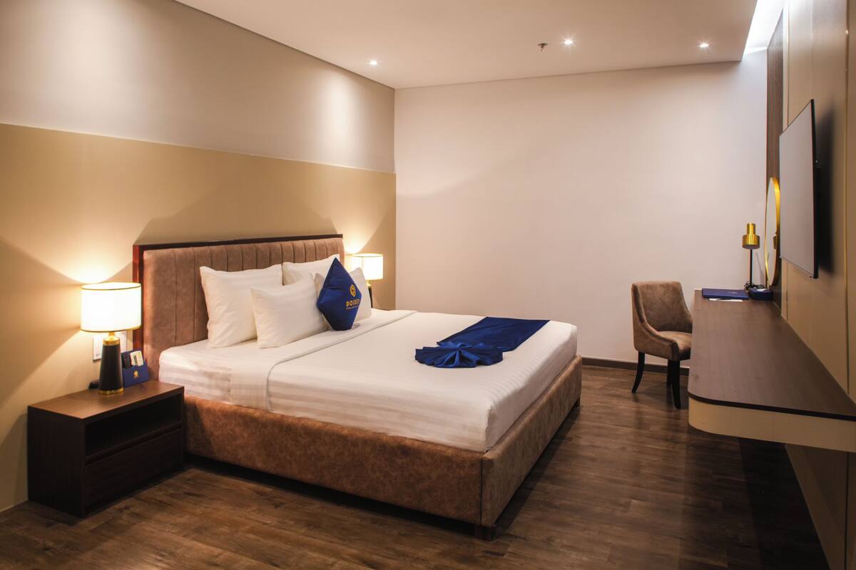 Quarto de hotel com uma cama grande, uma mesa de cabeceira com luminária, uma cadeira e uma escrivaninha com TV de tela plana.