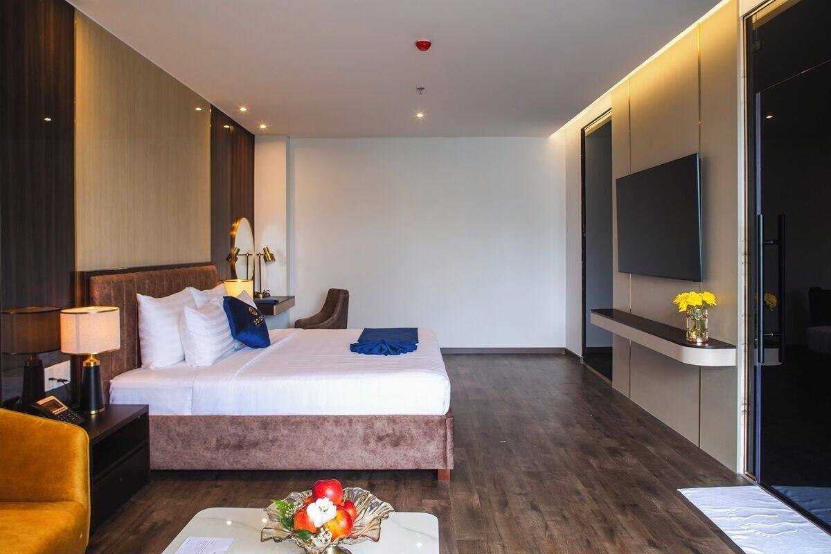 Quarto de hotel moderno com uma cama grande, mesas de cabeceira, televisão de tela plana e uma pequena mesa com frutas.
