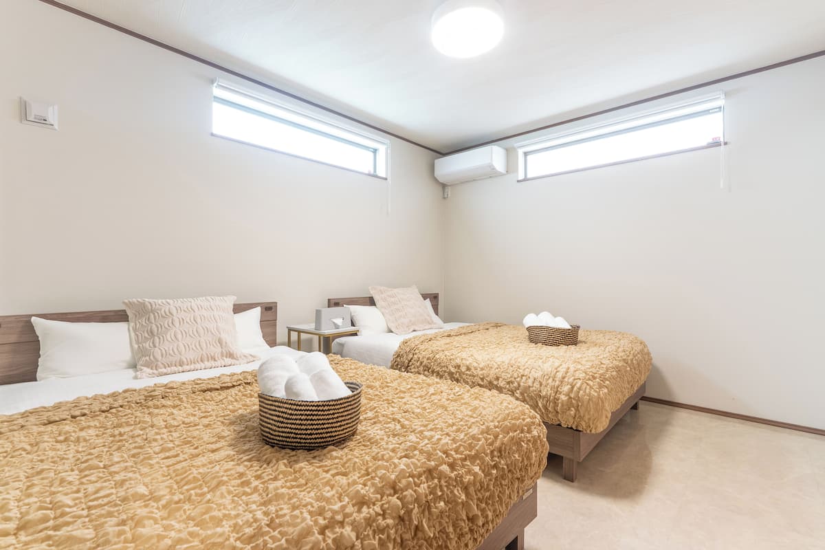 Quarto de hotel com duas camas, cada uma com um cobertor bege texturizado e uma cesta de vime. Há duas mesas de cabeceira com luminárias, e o quarto tem um teto branco com luminária redonda.