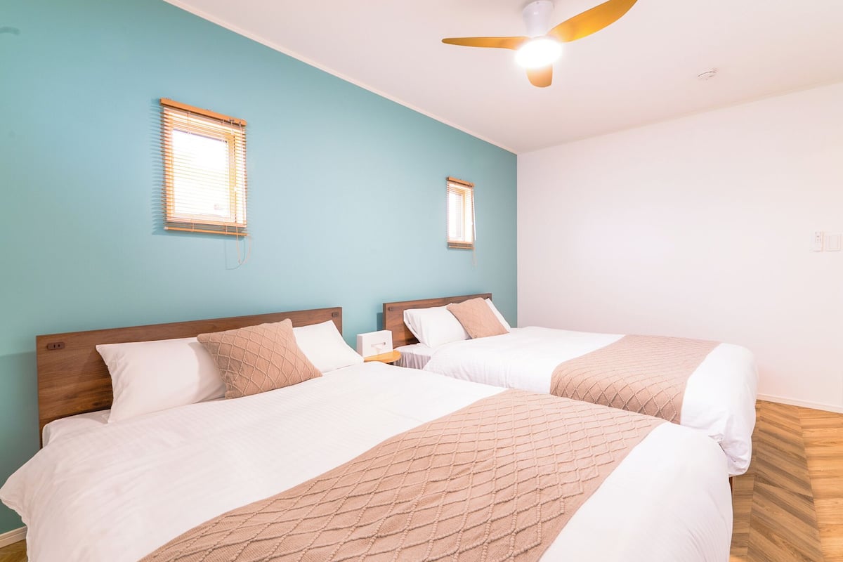 Quarto com duas camas, um ventilador de teto e duas janelas com persianas.