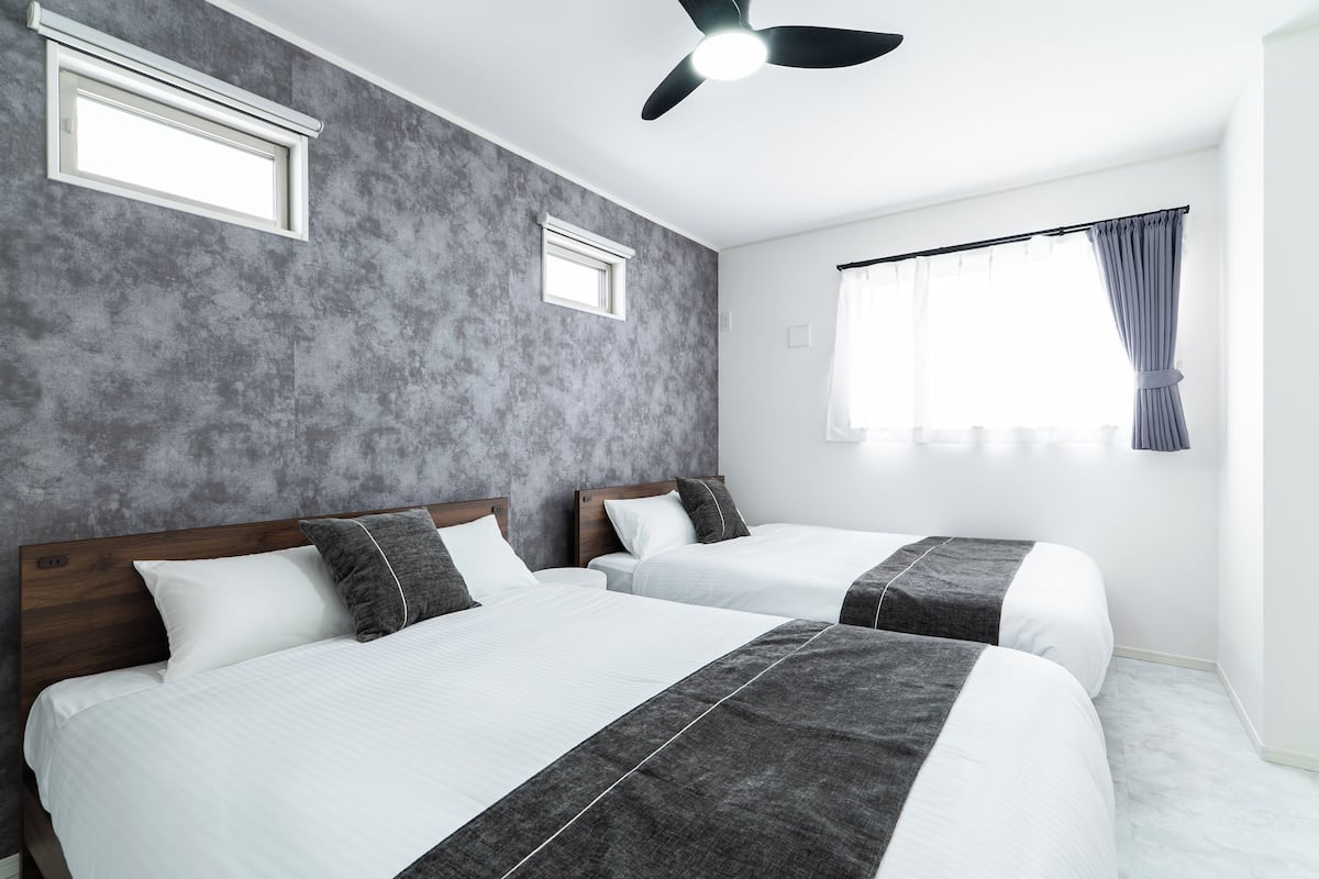 Quarto de hotel com duas camas, um ventilador de teto, uma janela com cortinas e uma parede cinza com textura.