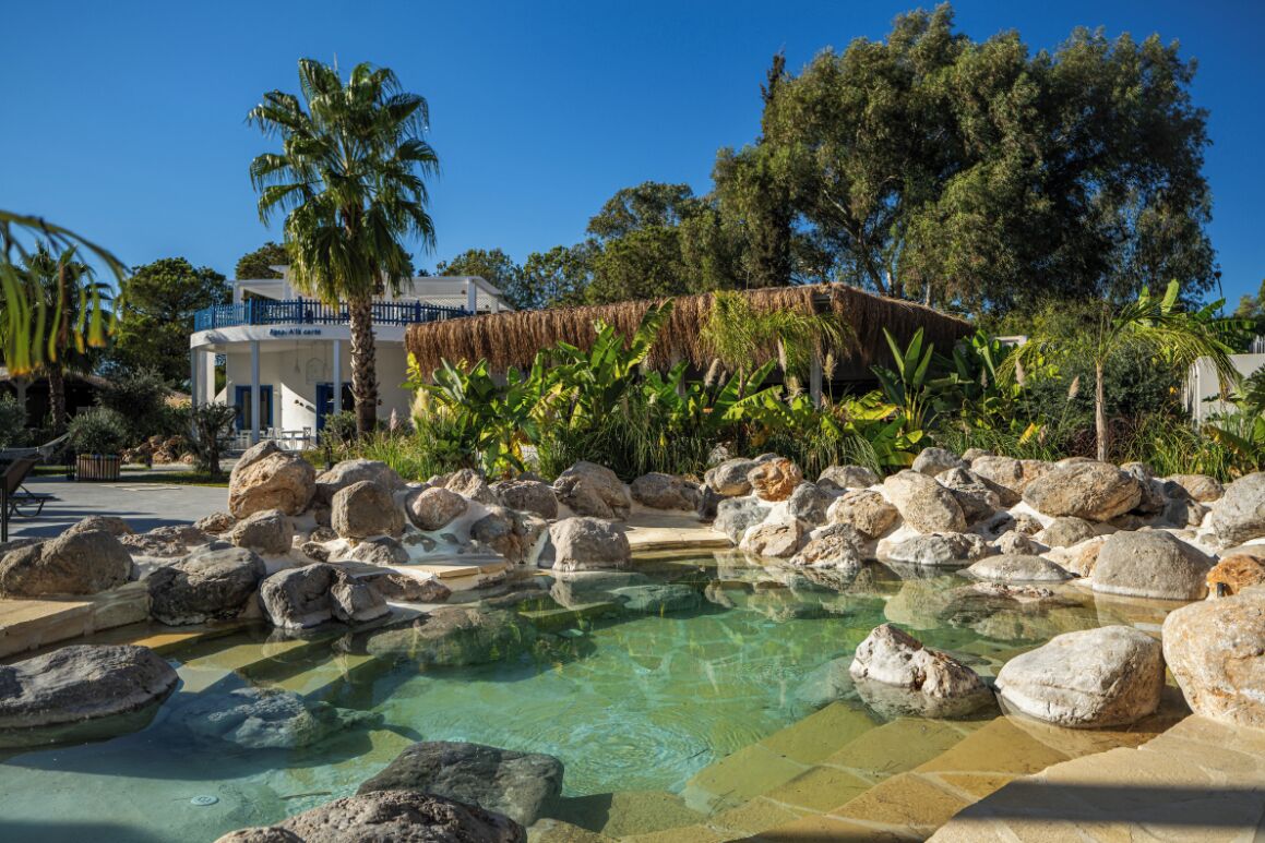 Uma piscina cristalina rodeada por pedras e plantas tropicais, com um edifício ao fundo.
