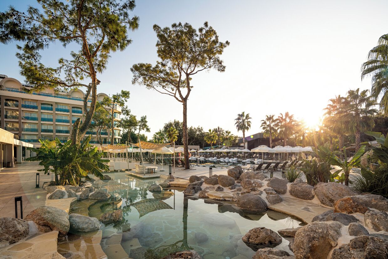 Área de piscina em resort, com um elemento de água cristalina, rodeada por rochas e palmeiras, e um edifício com varandas ao fundo.