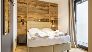 Quarto de hotel moderno com parede de painel de madeira, uma cama grande com toalhas dobradas e lustres laterais, e vista para o exterior por uma janela ampla.
