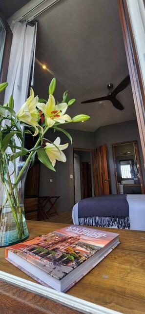 Quarto com cama, ventilador de teto, janela com cortinas e mesa com um livro e um vaso de flores.