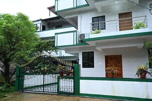Um edifício residencial de vários andares com fachada branca e detalhes verdes, apresentando uma entrada com portão e vasos de plantas.