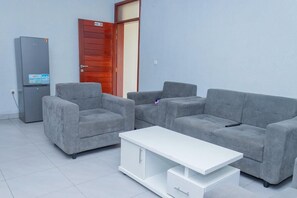 Quarto com sofá cinzento, mesa de centro branca e frigorífico cinzento.