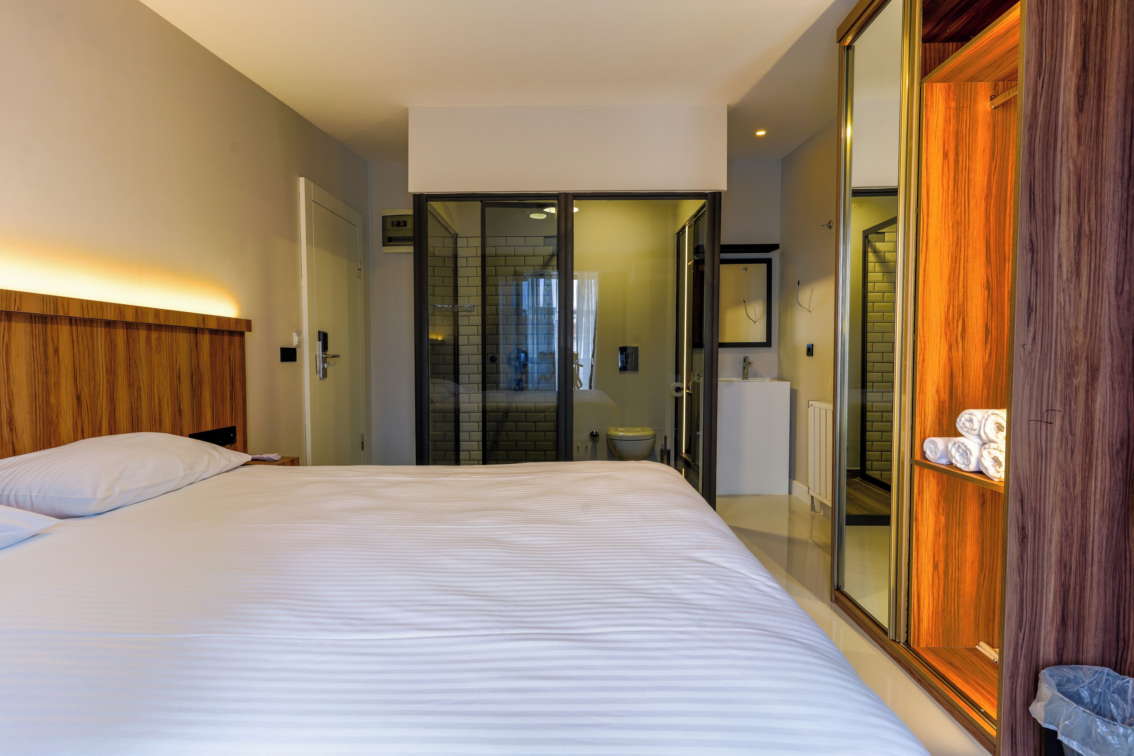 Quarto de hotel moderno com uma cama grande, um guarda-roupa de madeira, uma cabine de chuveiro de vidro e vista para o banheiro.