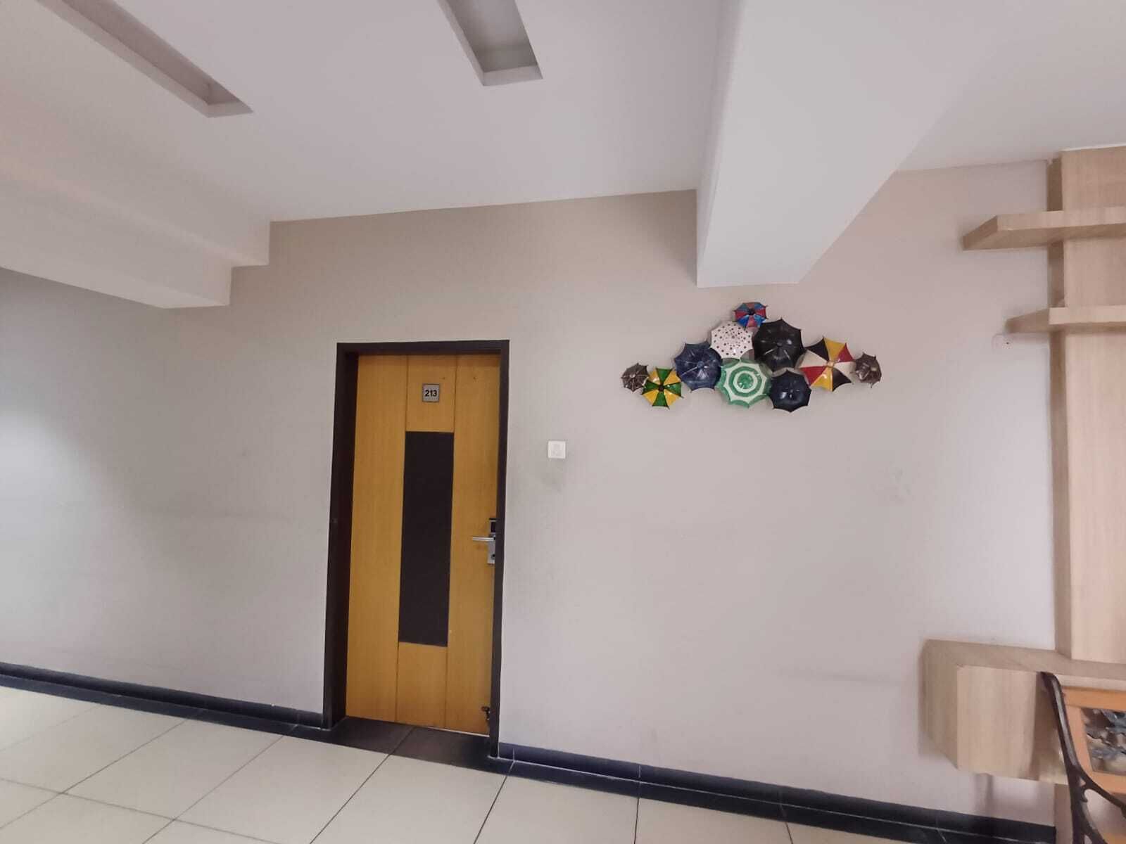 Corredor com porta de madeira marcada com o número 213, chão ladrilhado e parede com coleção de guarda-chuvas coloridos.