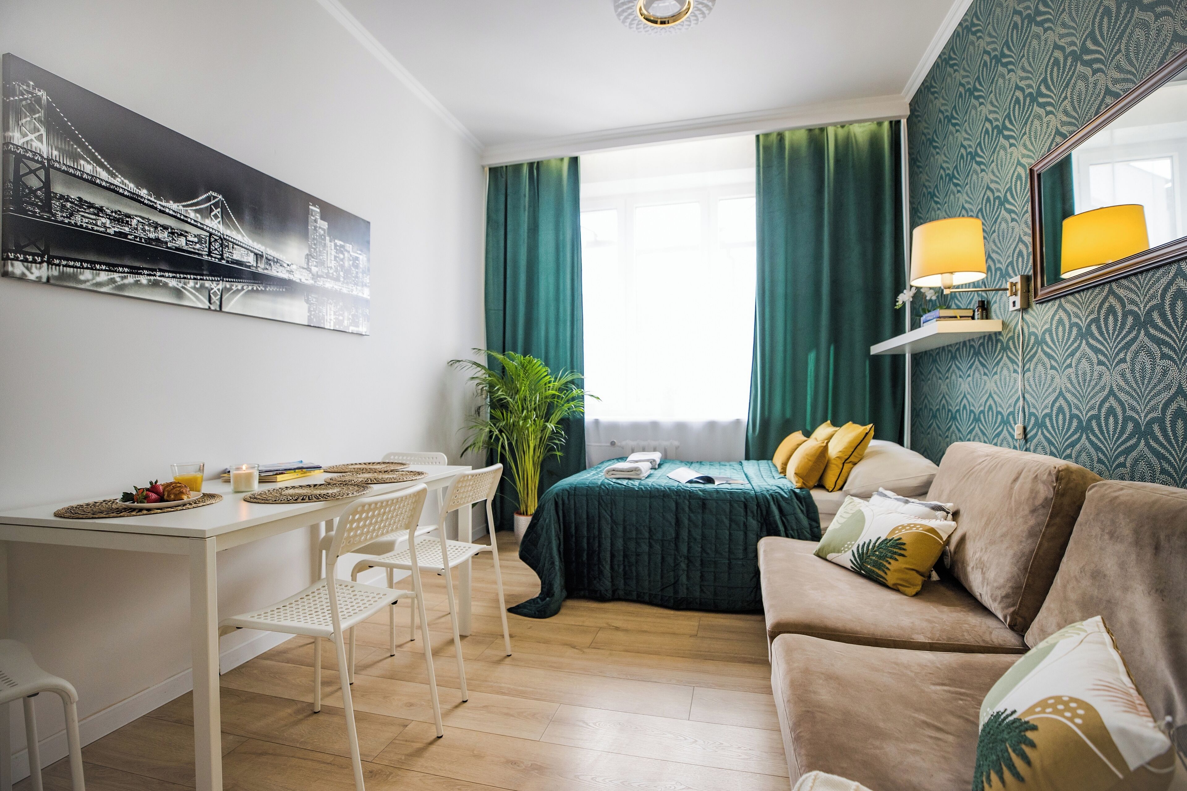 Quarto com cama, sofá, escrivaninha com cadeiras, mesa com planta e quadro na parede.