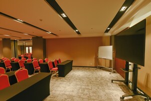 Uma sala de conferência com fileiras de cadeiras vermelhas, uma mesa preta, uma tela grande e um quadro branco.