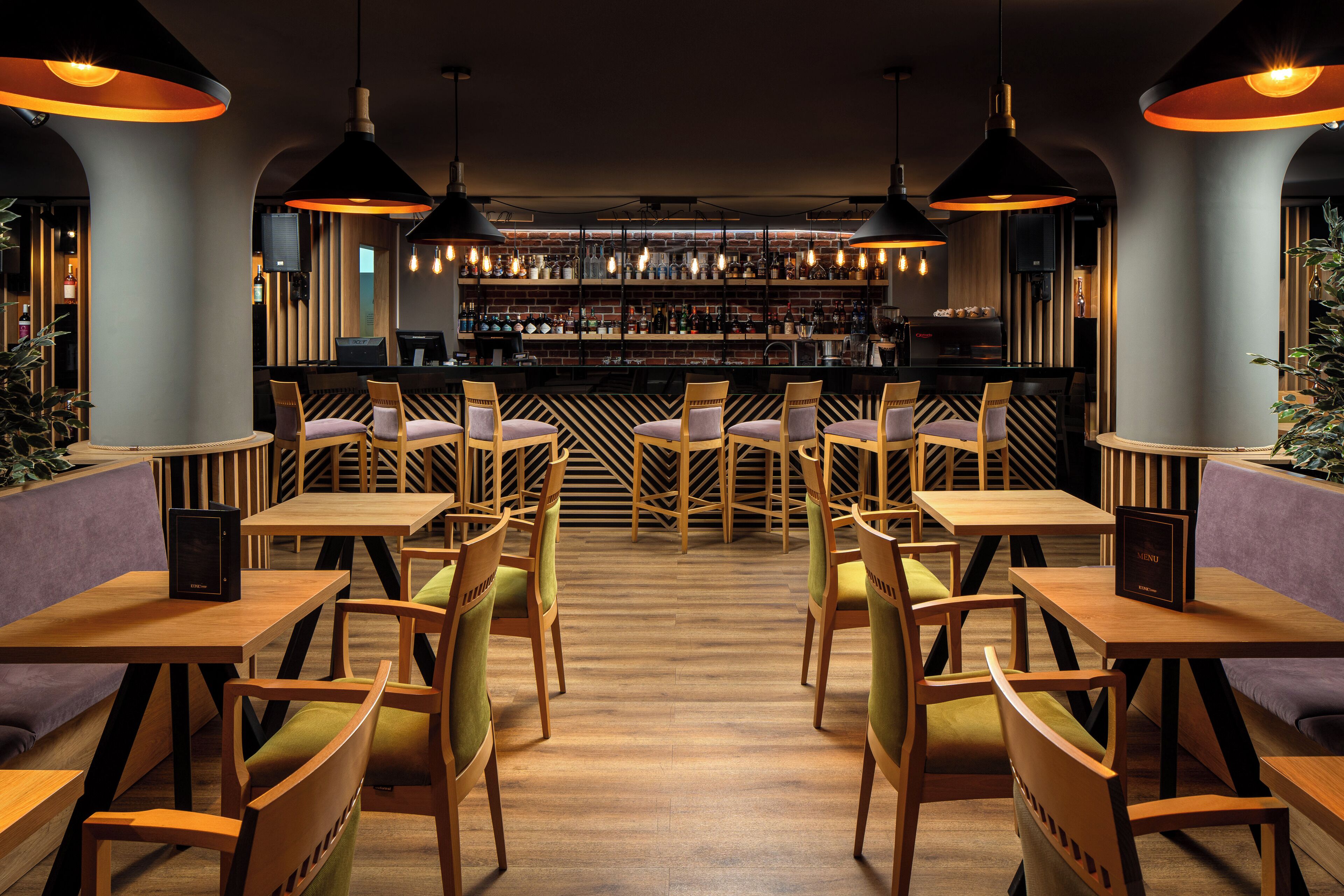 Restaurante com mesas e cadeiras de madeira, um bar ao fundo e luminárias pendentes.