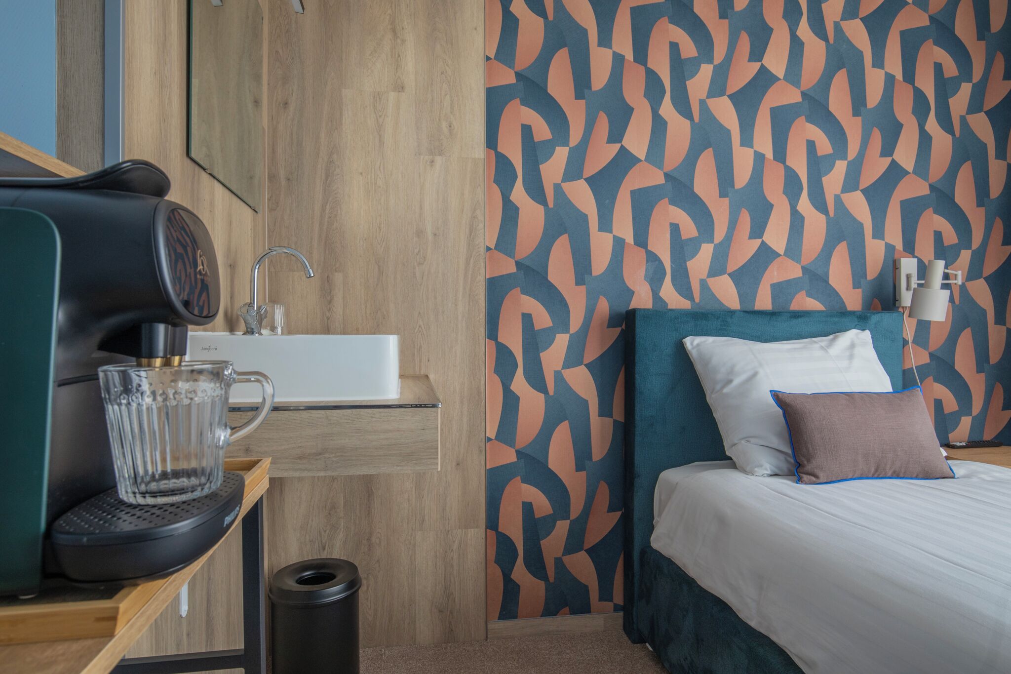Quarto de hotel com uma cama, uma mesinha de cabeceira com cafeteira, pia e parede estampada.