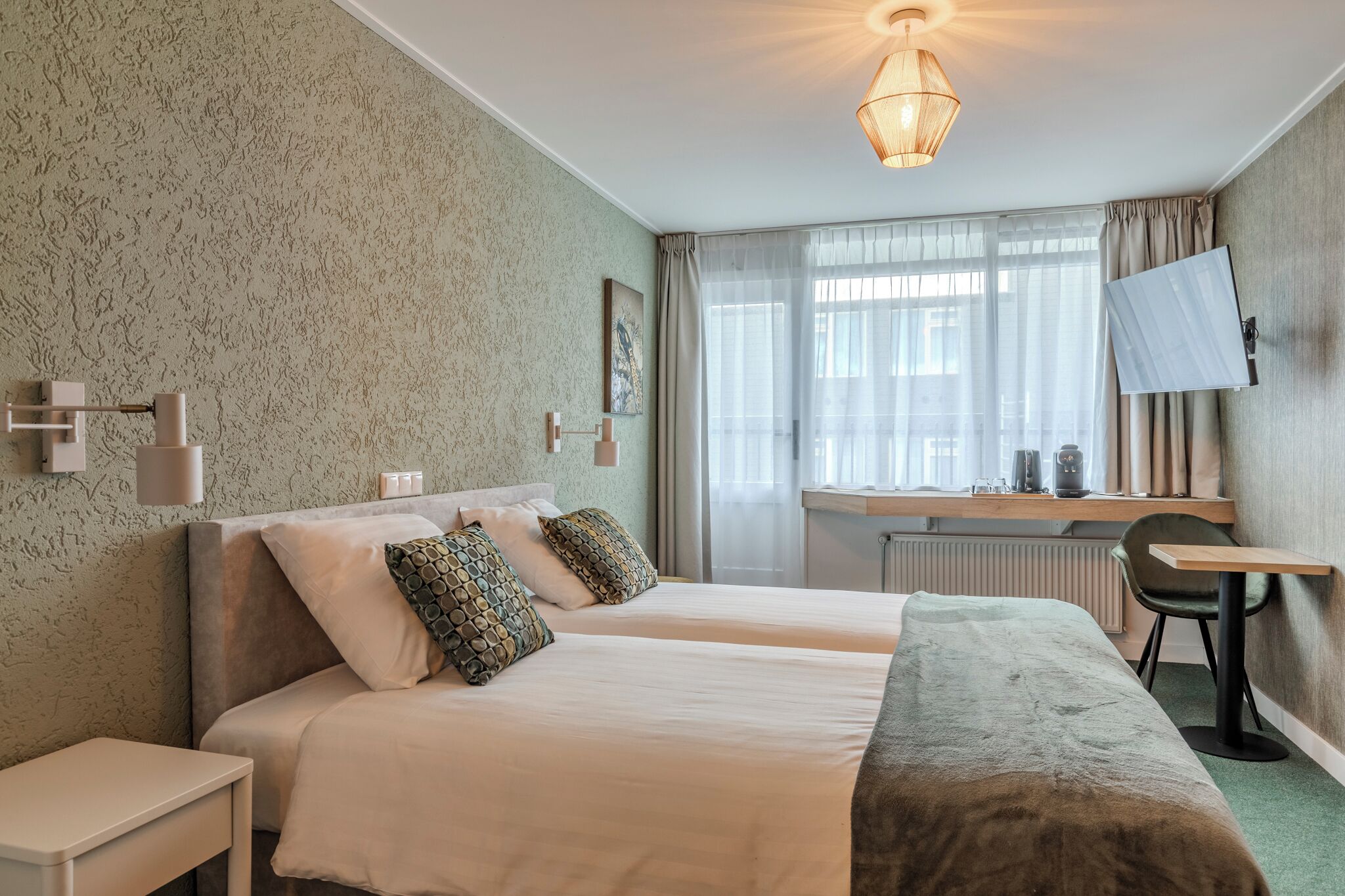 Quarto de hotel com duas camas, uma escrivaninha, uma cadeira, uma televisão e uma janela com cortinas.