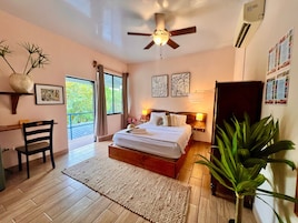 Quarto com cama, escrivaninha, cadeira, ventilador de teto e uma janela grande com cortinas.