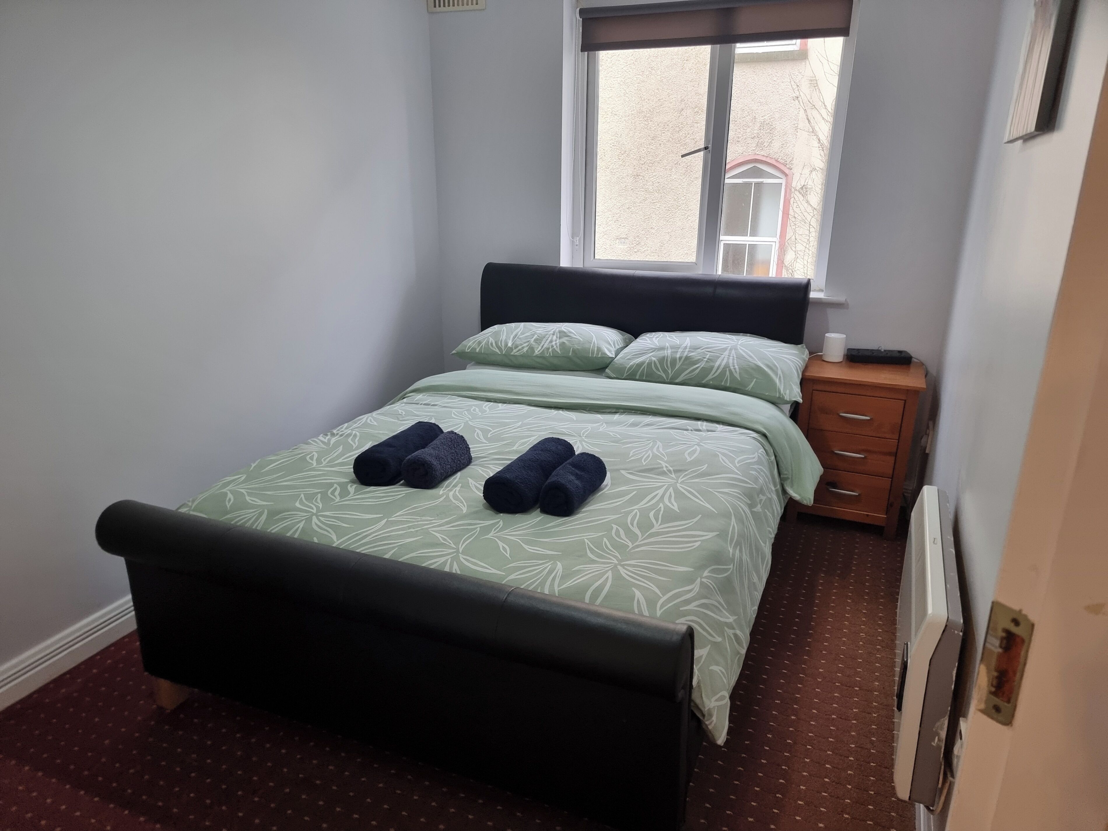 Cama bem arrumada com colcha e almofadas estampadas em verde, mesa de cabeceira de madeira com abajur e janela com vista para um edifício.