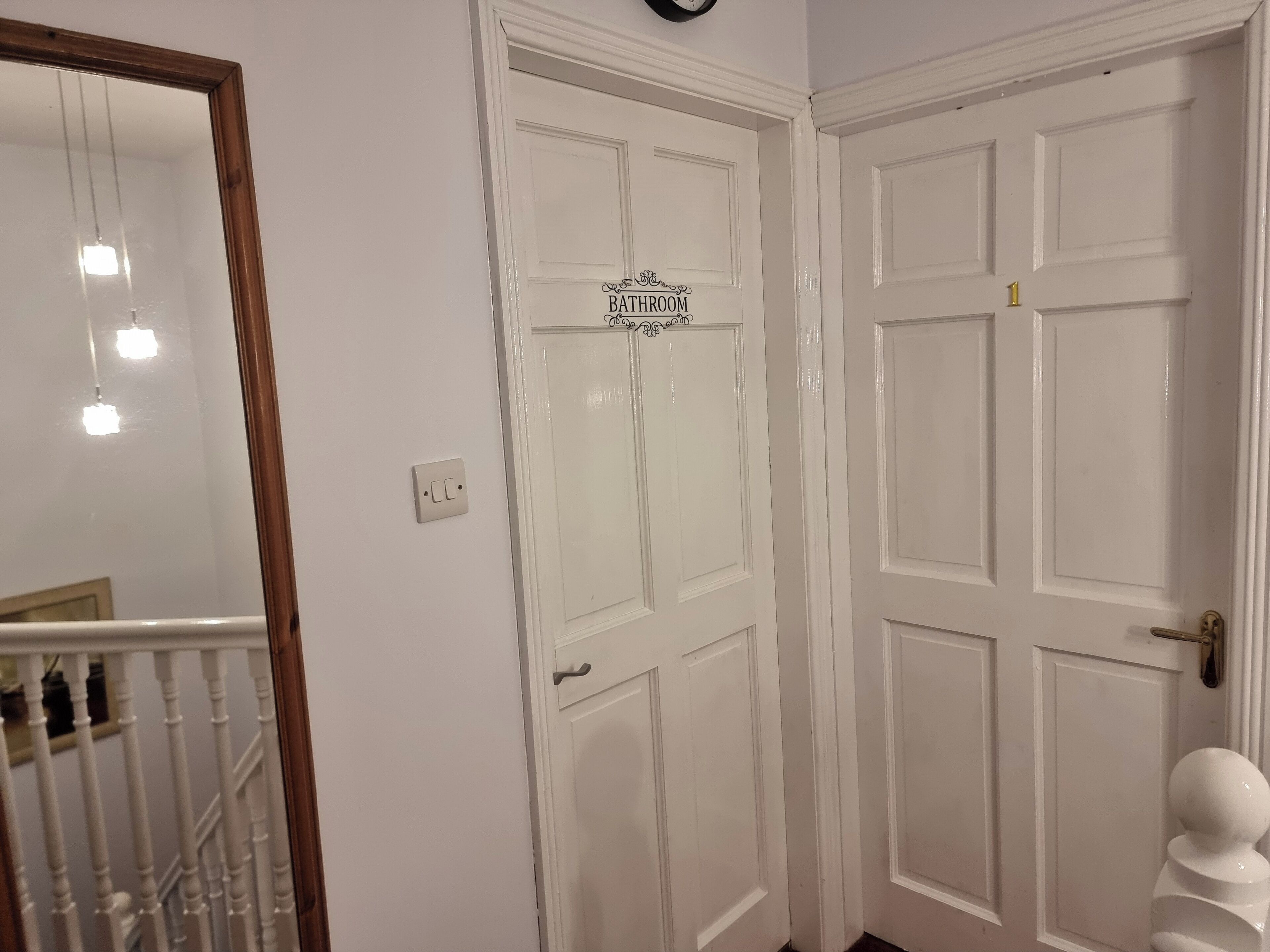 Uma porta branca com a placa 'Bathroom', uma parede branca e um espelho com moldura de madeira.