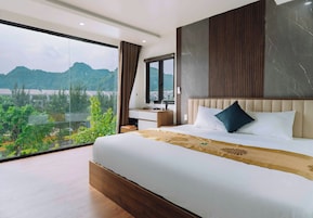 Quarto de hotel moderno com uma cama grande, mesa de cabeceira de madeira, escrivaninha e uma janela ampla com vista para vegetação e montanhas.