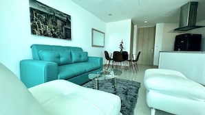 Sala de estar moderna com um sofá azul-turquesa, um sofá branco e uma mesa de centro de vidro. Há uma área de jantar com mesa e cadeiras, e uma cozinha com fogão e geladeira.