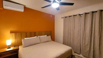 Quarto de hotel com uma cama, mesa de cabeceira, ar condicionado, ventoinha no teto e cortinas.