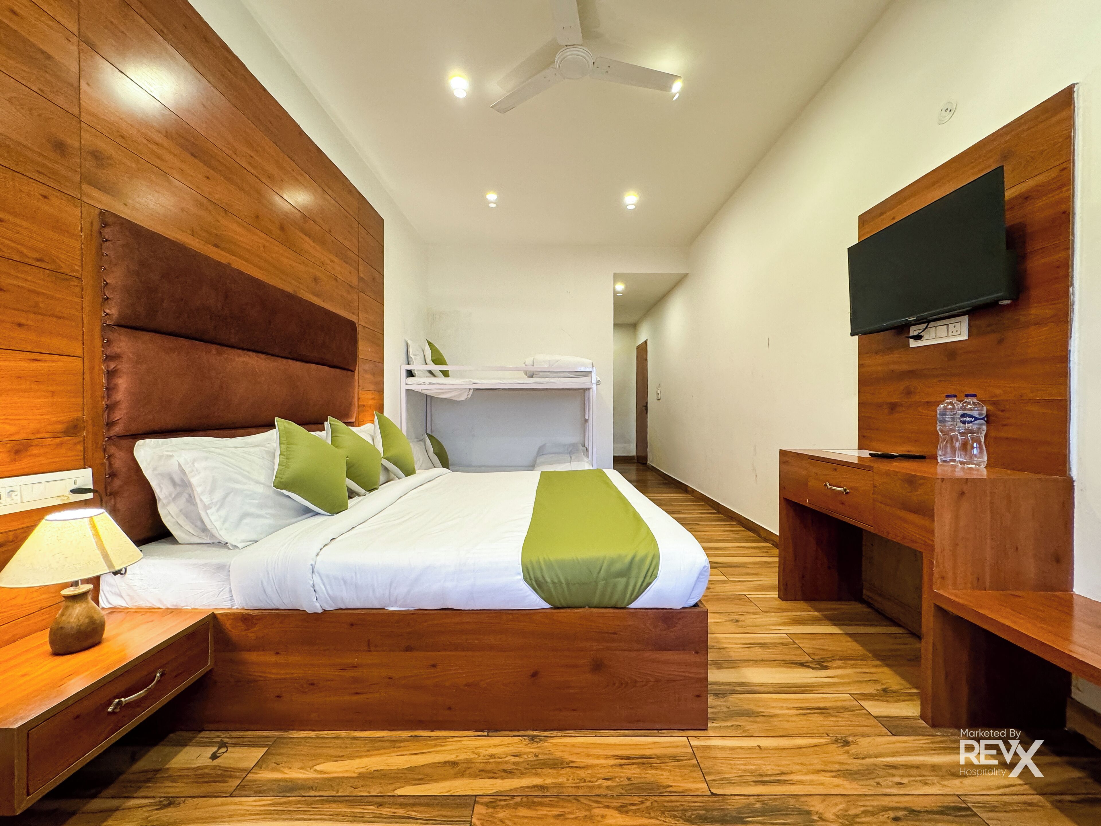 Quarto de hotel com cabeceira de madeira, cama com roupa de cama verde e branca, mesa de cabeceira com abajur, escrivaninha de madeira, televisão de tela plana e dispensador de água.