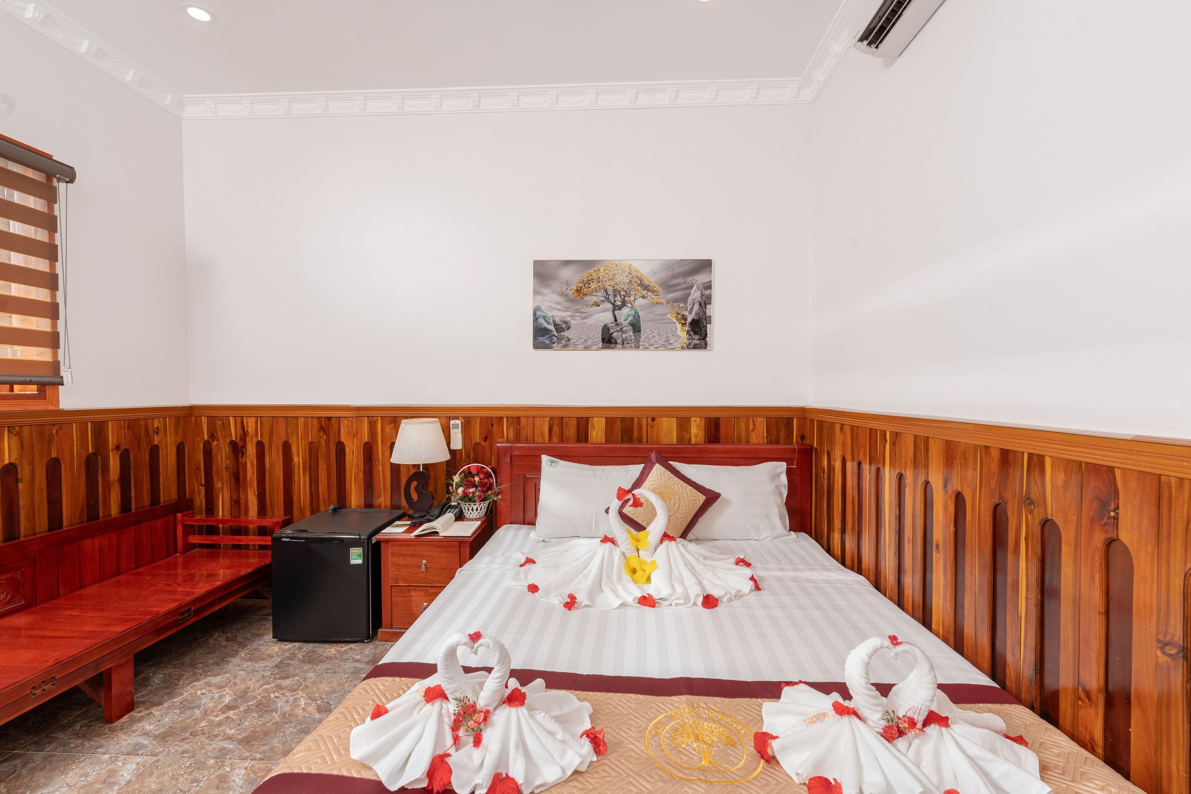 Quarto de hotel com cabeceira de madeira, cama com roupa de cama branca, um banquinho vermelho, um refrigerador preto e um quadro na parede.