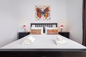 Uma cama de casal bem arrumada com lençóis brancos, dois travesseiros e duas mesas de cabeceira com luminárias.