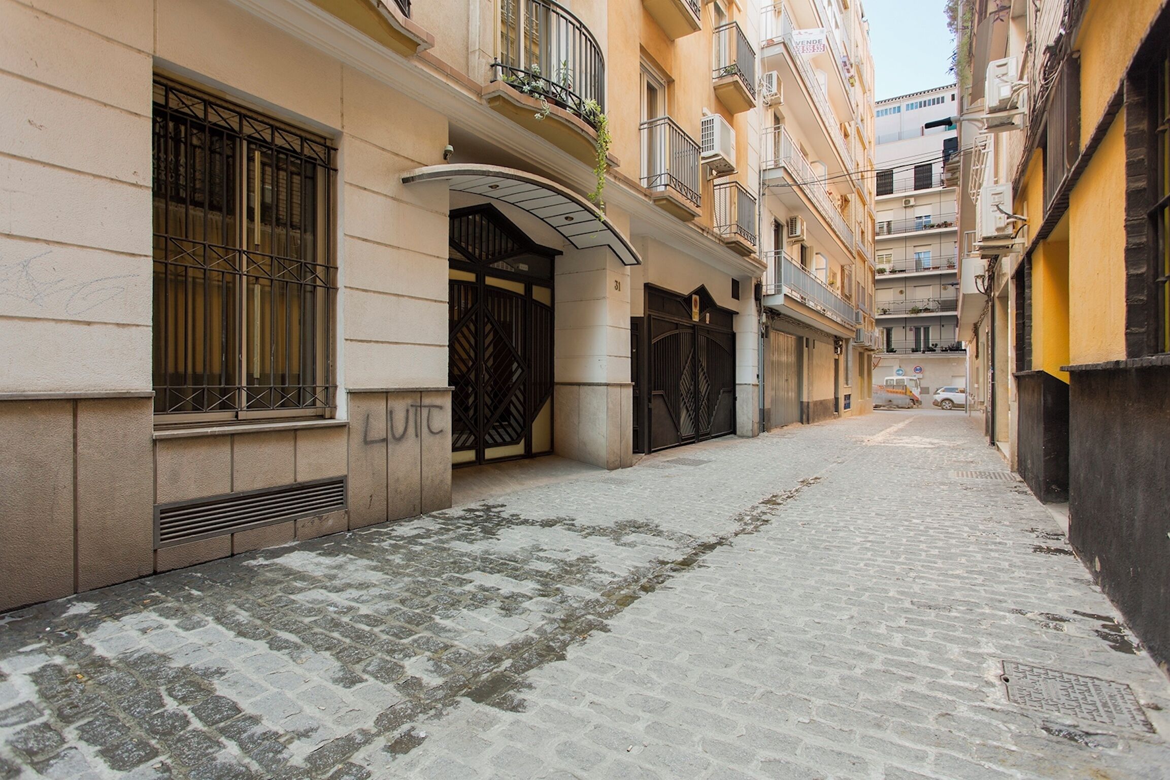 Uma rua de paralelepípedos estreita, ladeada por edifícios altos com janelas protegidas por grades e varandas.