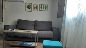 Uma sala de estar com um sofá cinza, uma mesa de centro de madeira e um banquinho azul-esverdeado. Há impressões emolduradas de folhas na parede.