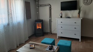 Um quarto aconchegante com lareira a lenha, televisão fixada na parede, cômoda branca e janela com cortinas leves.