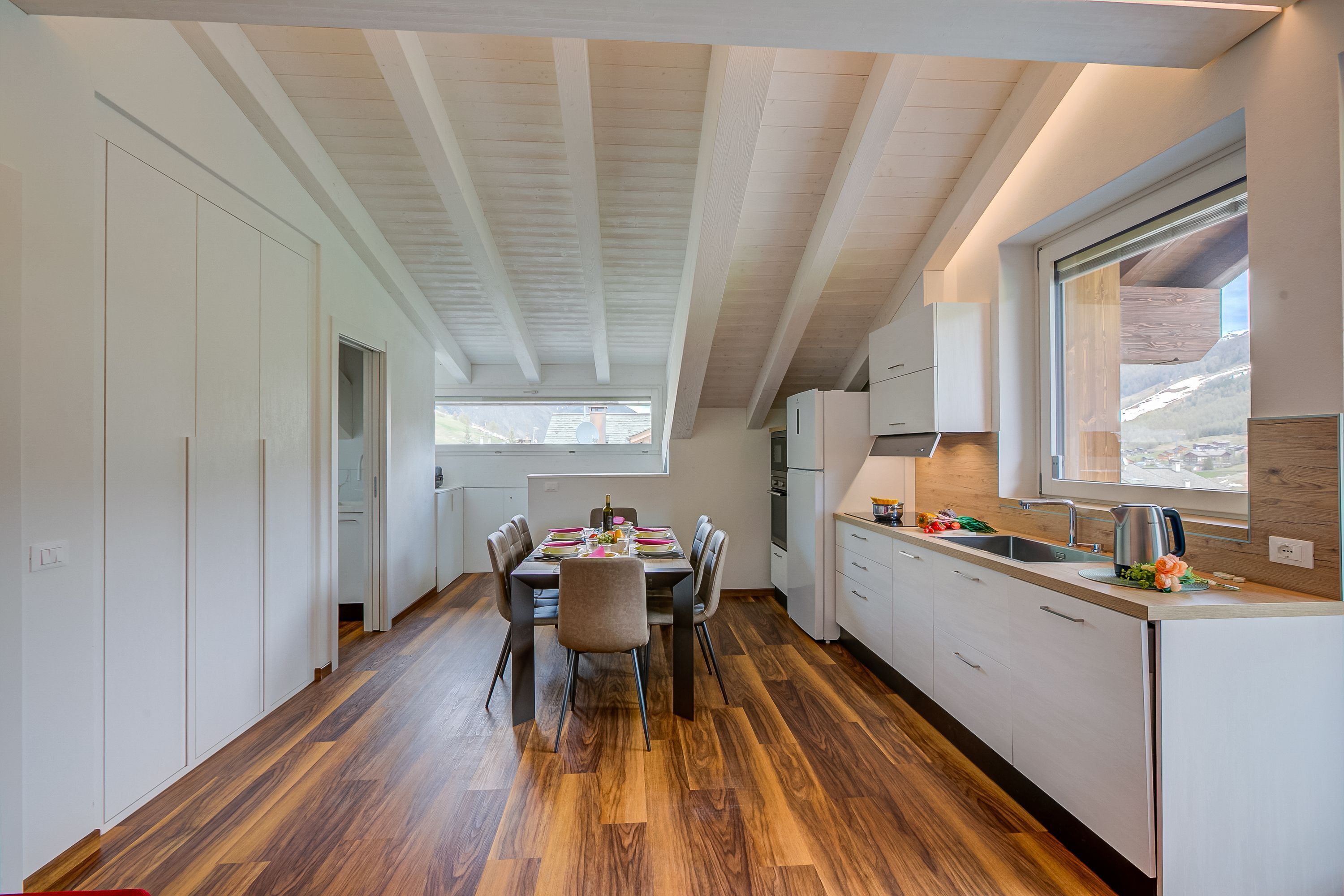 Uma cozinha moderna com piso de madeira, armários brancos e um espaço de refeições com mesa posta para quatro pessoas.