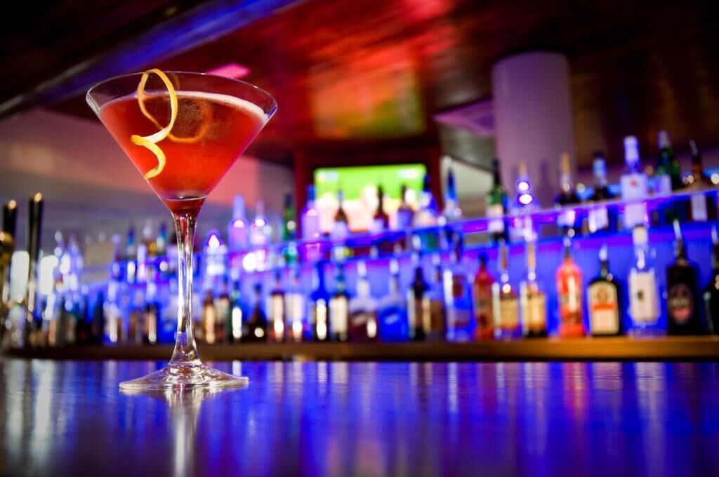 Um copo de martini com uma rodela de limão sobre um balcão de bar, com várias garrafas ao fundo.