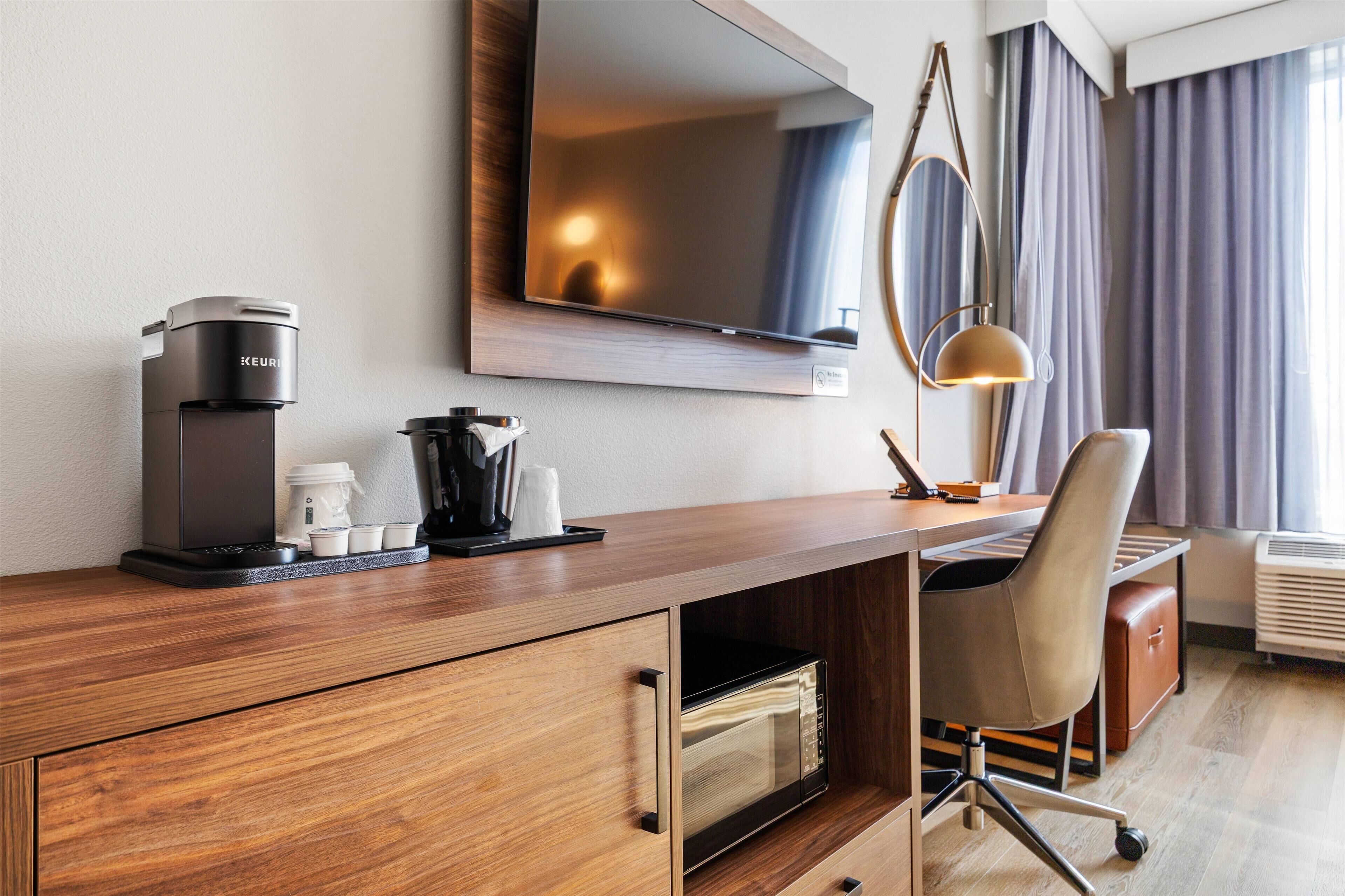 Quarto de hotel moderno com uma escrivaninha de madeira, uma cafeteira Keurig, micro-ondas, um espelho grande e uma luminária.