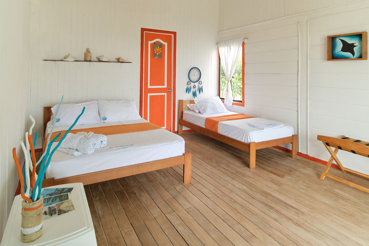 Quarto com duas camas de madeira, um banquinho de madeira, uma estante com objetos decorativos e um sonho na parede.