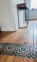 Quarto com chão de madeira, tapete estampado e mesa de metal preta em frente a uma parede branca.