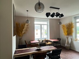 Uma sala de jantar com uma mesa de madeira, cadeiras e dois vasos grandes com plumas de capim-do-campo. Há luminárias pendentes e um candeeiro no teto.
