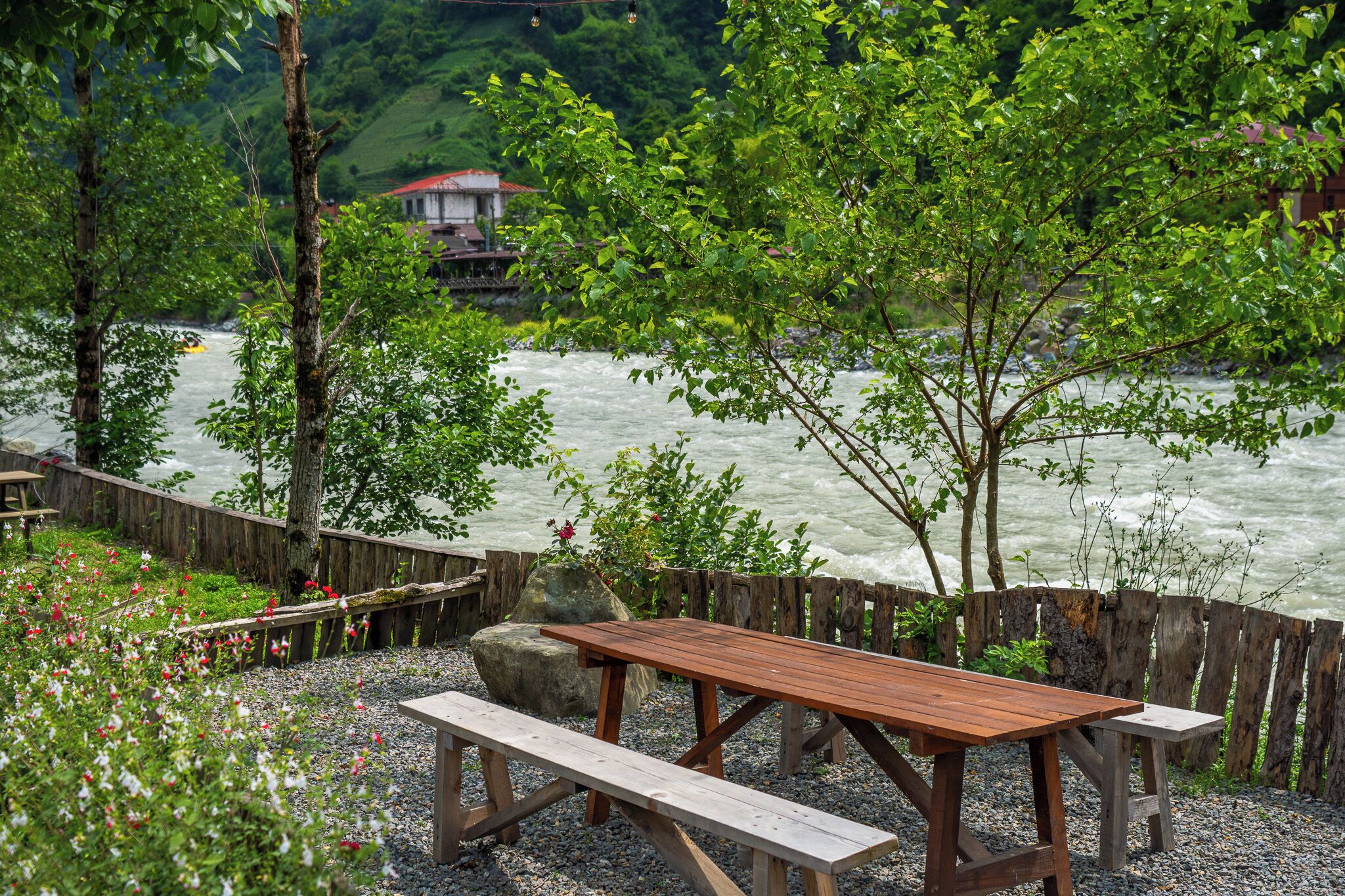 Mesa de piquenique de madeira e bancos à beira de um rio, com cerca de madeira e vegetação.