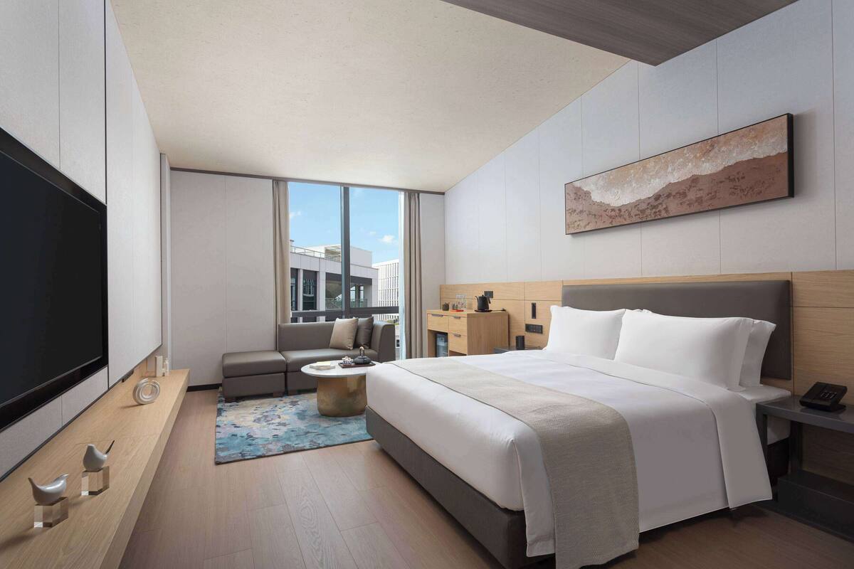 Quarto de hotel moderno com uma cama grande, televisão de tela plana, sofá, mesa de centro e vista para a cidade.