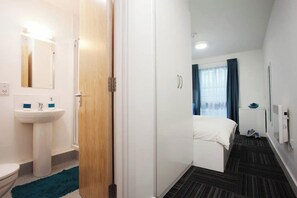 Quarto de hotel com pia branca, espelho, cama com colcha branca e chão coberto por carpete escuro.