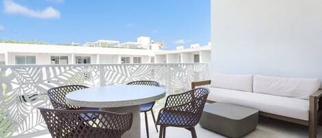 Varanda com mesa redonda e cadeiras, um sofá branco e vista para edifícios ao fundo.