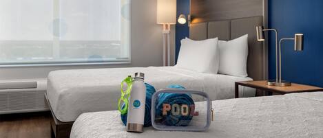Quarto de hotel com duas camas, janela com persianas, luminária de cabeceira, garrafa de água, um par de óculos e uma bola azul com a palavra 'POOL' escrita.
