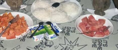 Mesa posta ao ar livre com diversos pratos, incluindo arroz, legumes e bebidas enlatadas, coberta por uma toalha com padrão de jogo.
