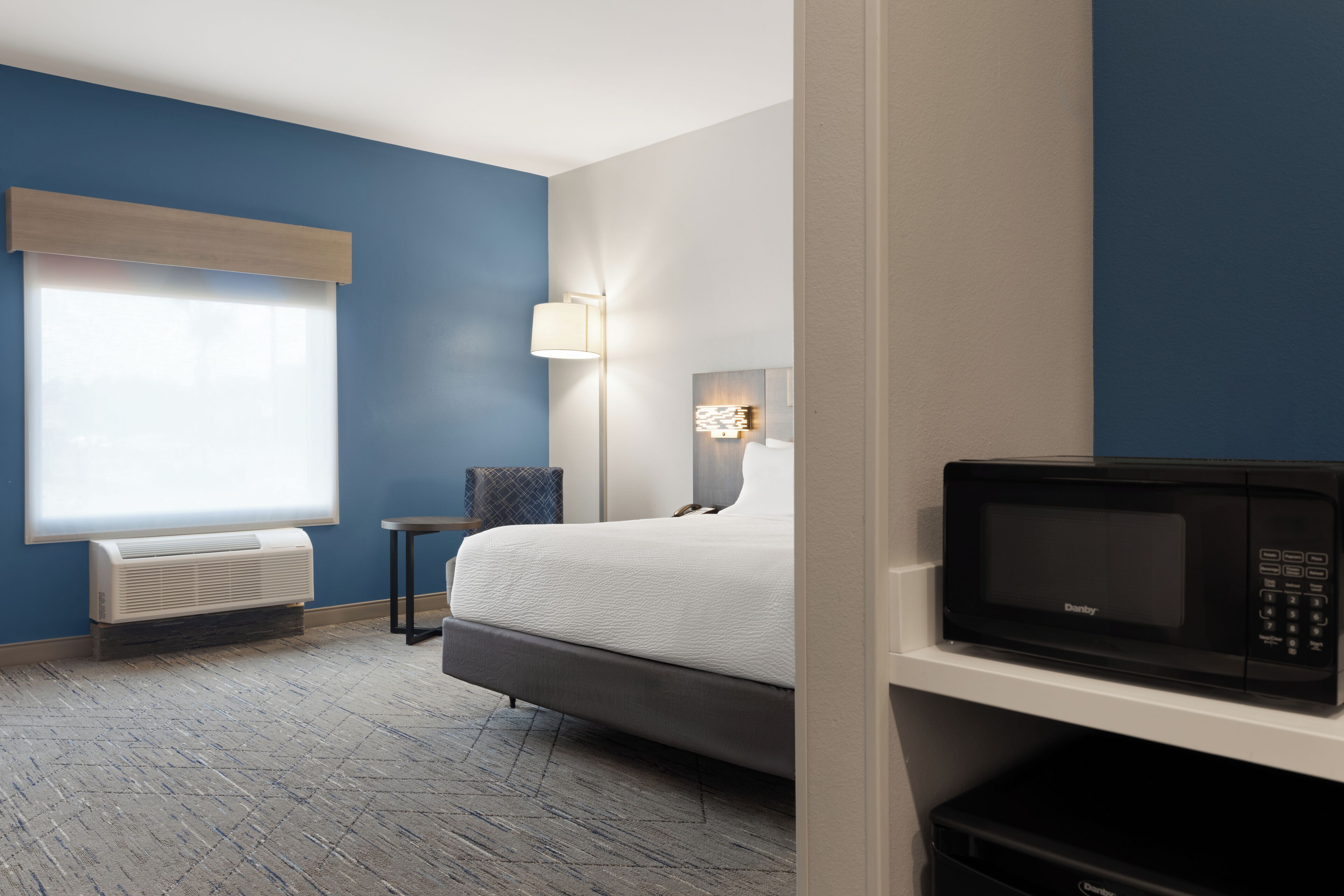 Quarto de hotel com cama, escrivaninha, cadeira, janela com persianas, luminária na parede e micro-ondas.