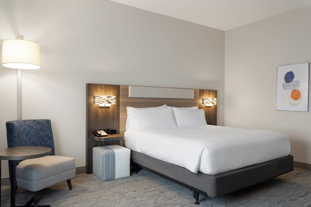 Quarto de hotel com cama, uma cadeira, mesa de cabeceira e abajur.