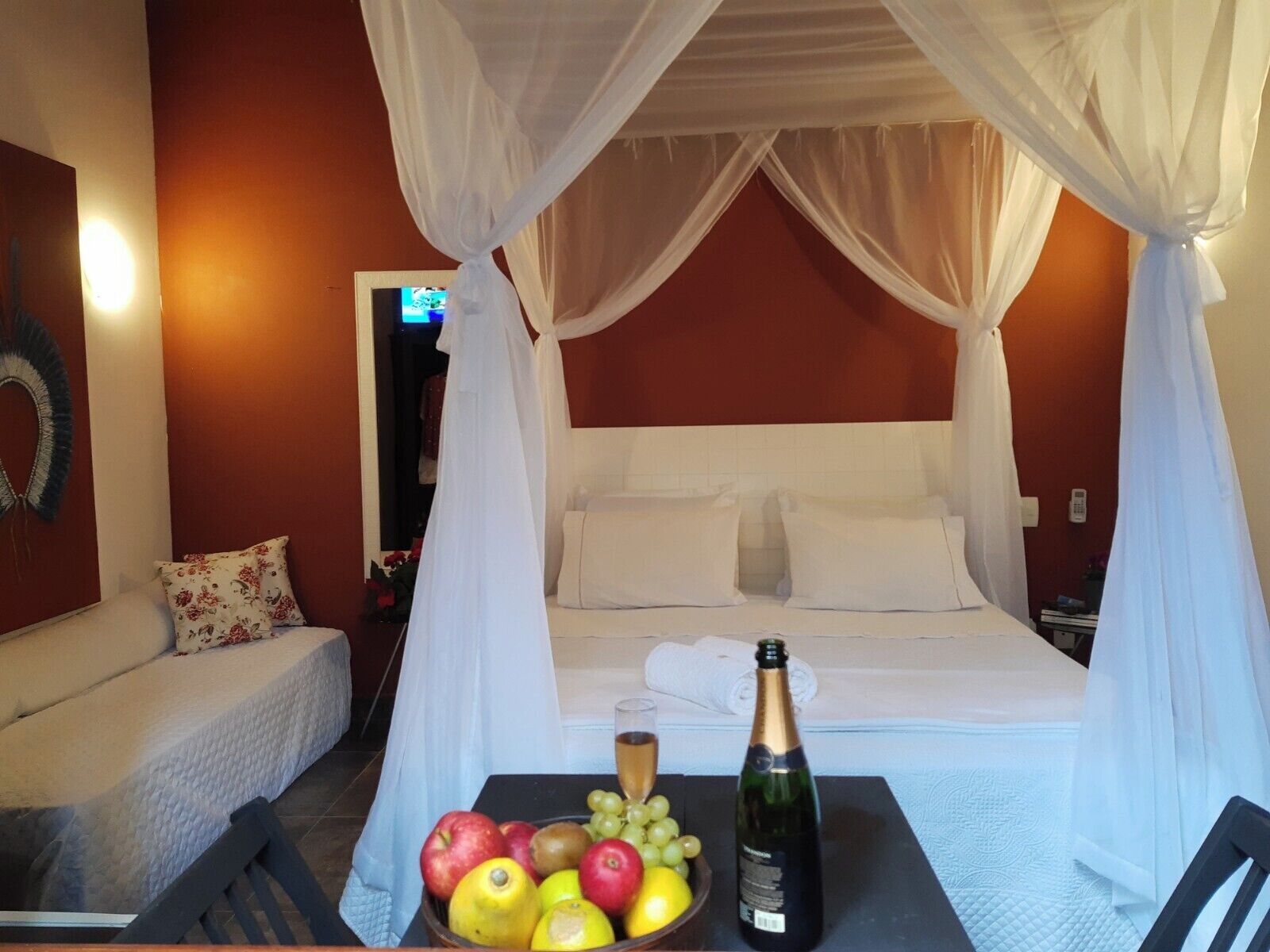Quarto com cama de dossel, mesa de cabeceira com uma garrafa de champanhe e uma cesta de frutas.
