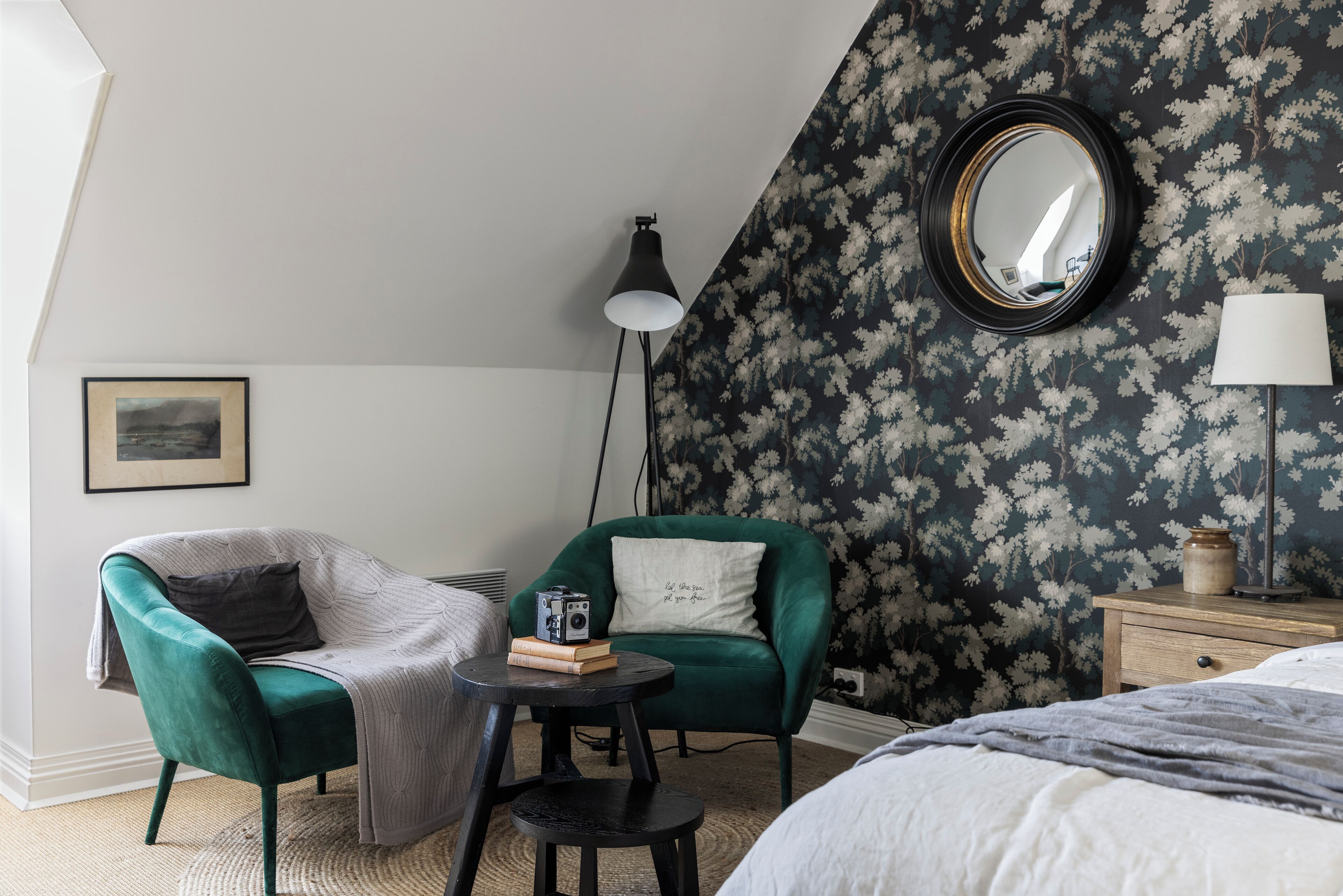 Quarto com uma cama, duas poltronas verdes, uma mesa pequena com livros, um abajur e um espelho na parede.