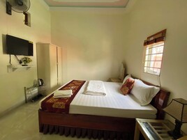 Quarto de hotel com cama, televisão fixada na parede, janela com persianas, ventilador e pequena mesa com abajur.