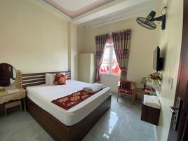 Quarto de hotel com uma cama grande, uma escrivaninha com espelho, uma cadeira, um guarda-roupa e uma pequena mesa com luminária e flores.