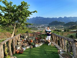 Uma pessoa está em pé numa varanda de madeira, com vista para um jardim exuberante, vários vasos com plantas e montanhas distantes, além de uma cidade.